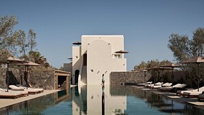 Uma área de piscina com espreguiçadeiras e guarda-sóis, uma parede de pedra e um edifício com telhado plano.