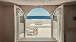 Quarto com uma grande janela em arco que dá para uma varanda com cadeiras de descanso e vista para o mar.
