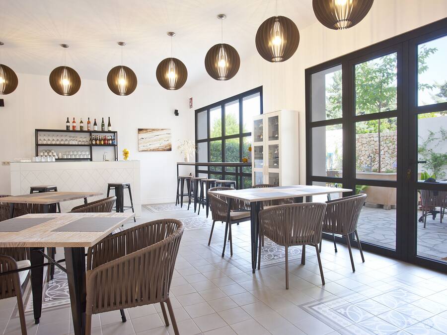 Restaurante com várias mesas e cadeiras, área de bar com garrafas e copos, e amplas janelas que oferecem vista para o exterior.