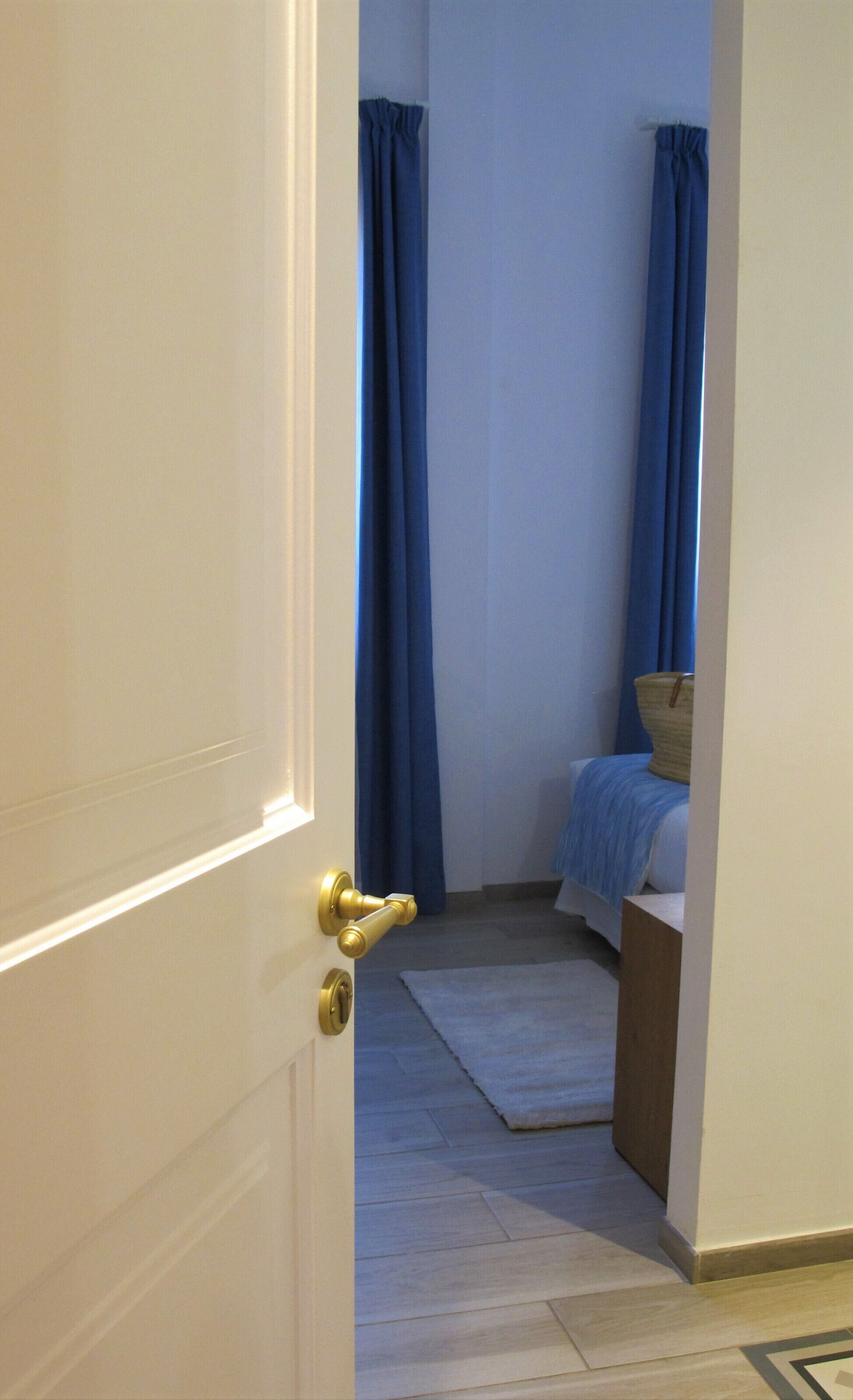 Um quarto com porta, cama, mesa de cabeceira e janela com cortinas azuis.