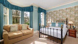 Quarto com cama, mesa de cabeceira de madeira, sofá bege e janela com cortinas.