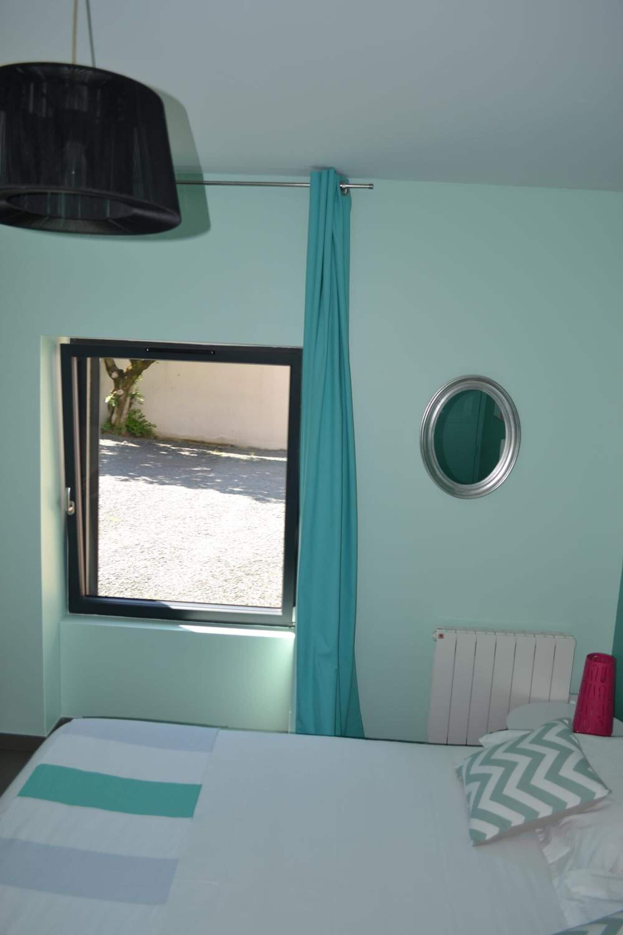 Um quarto com uma cama, janela com cortinas, radiador e um espelho na parede.