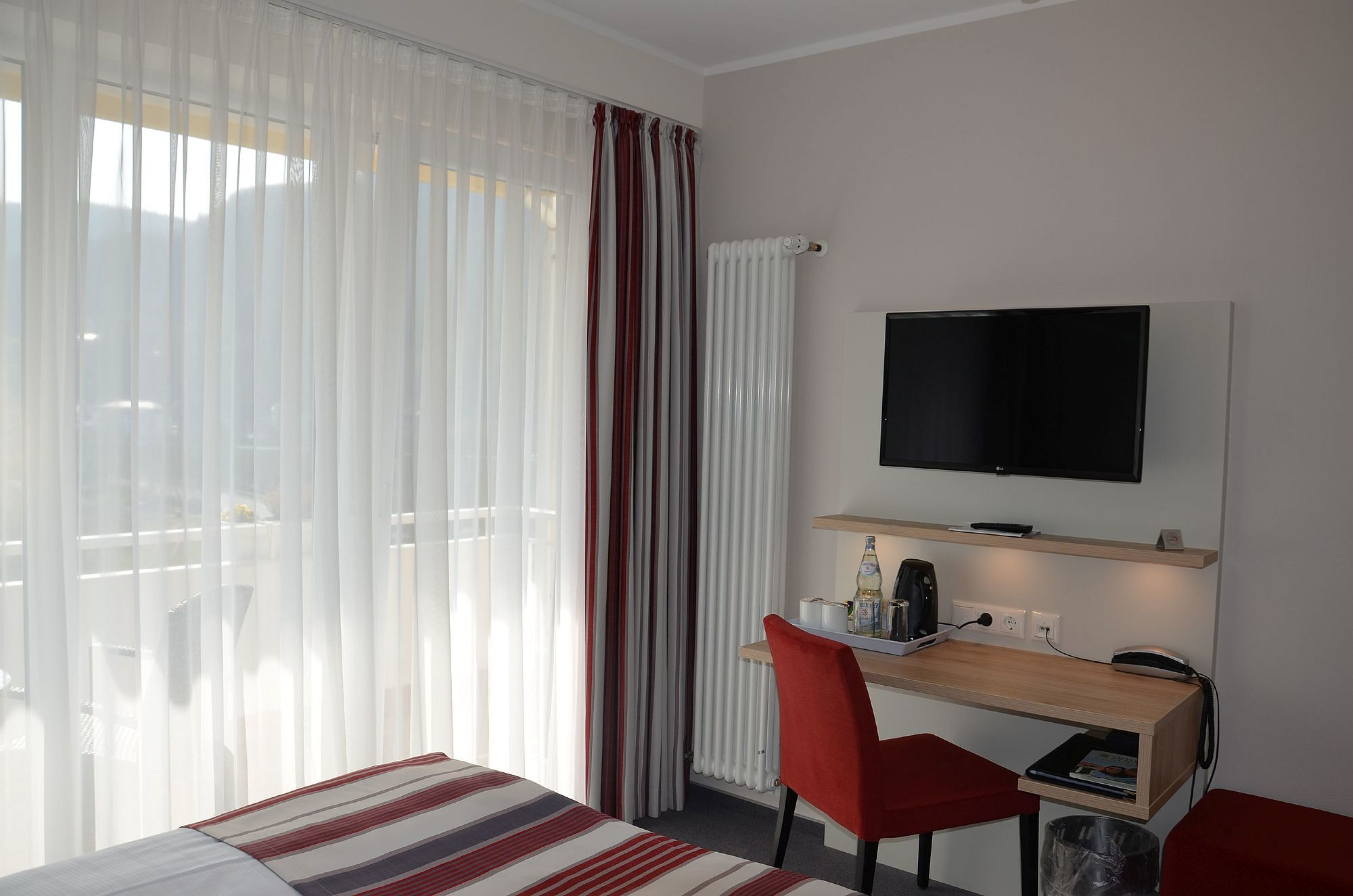 Quarto de hotel com uma cama, uma escrivaninha com cadeira, televisão e janela com cortinas.