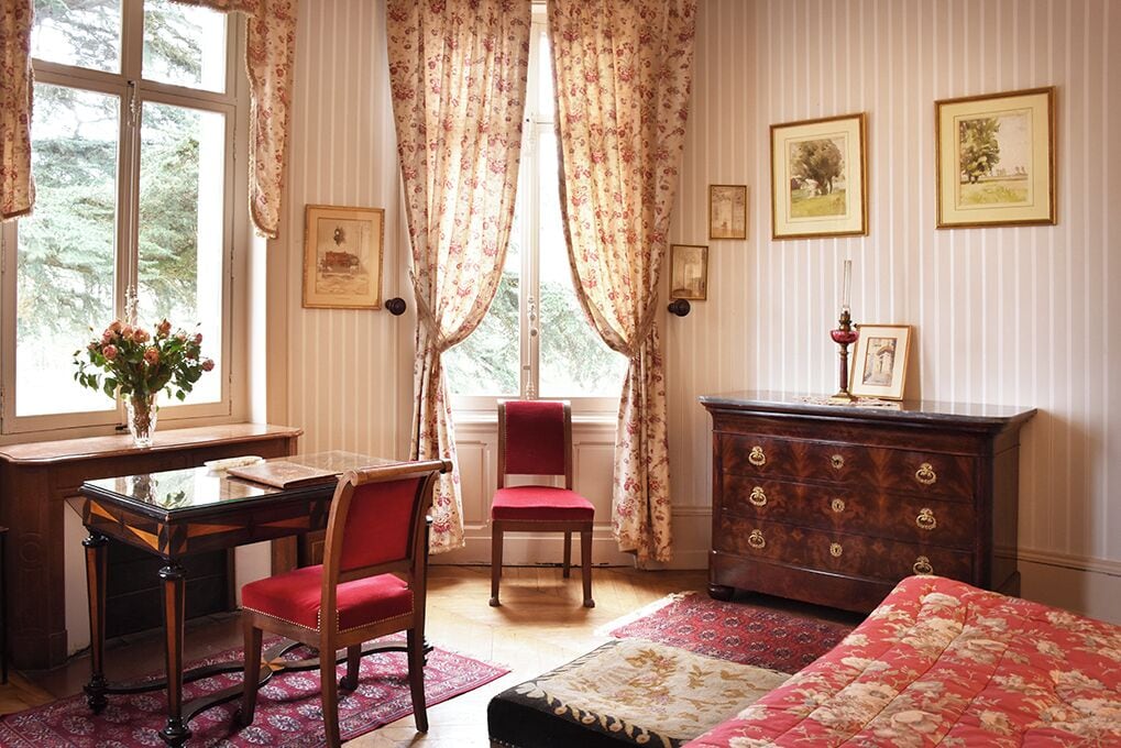 Quarto com escrivaninha de madeira, uma cadeira vermelha, cama com colcha florida, cômoda e cortinas. Há quadros nas paredes e um vaso com flores sobre a escrivaninha.