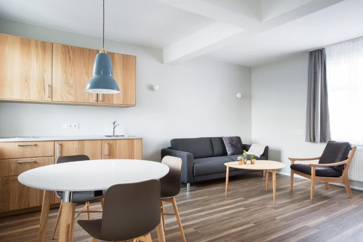 Uma sala de estar moderna com uma mesa de jantar, sofá e cadeiras. Há uma área de cozinha com armários de madeira e uma luminária pendente.