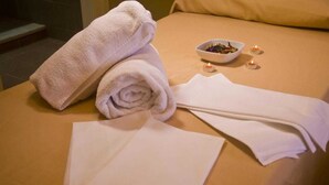 Cama bem arrumada com uma toalha branca enrolada, um recipiente com flores secas e duas velas acesas.