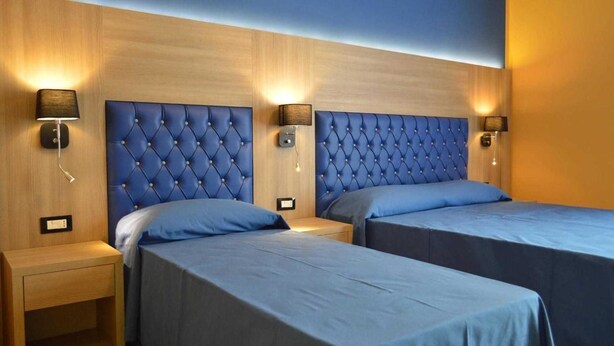 Quarto de hotel com duas camas, cada uma com cabeceira azul e almofadas combinando. Há mesas de cabeceira com abajures e uma luminária na parede.