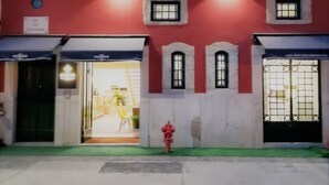 Um edifício vermelho com várias janelas e varandas. São visíveis duas portas, uma delas com uma placa acima.