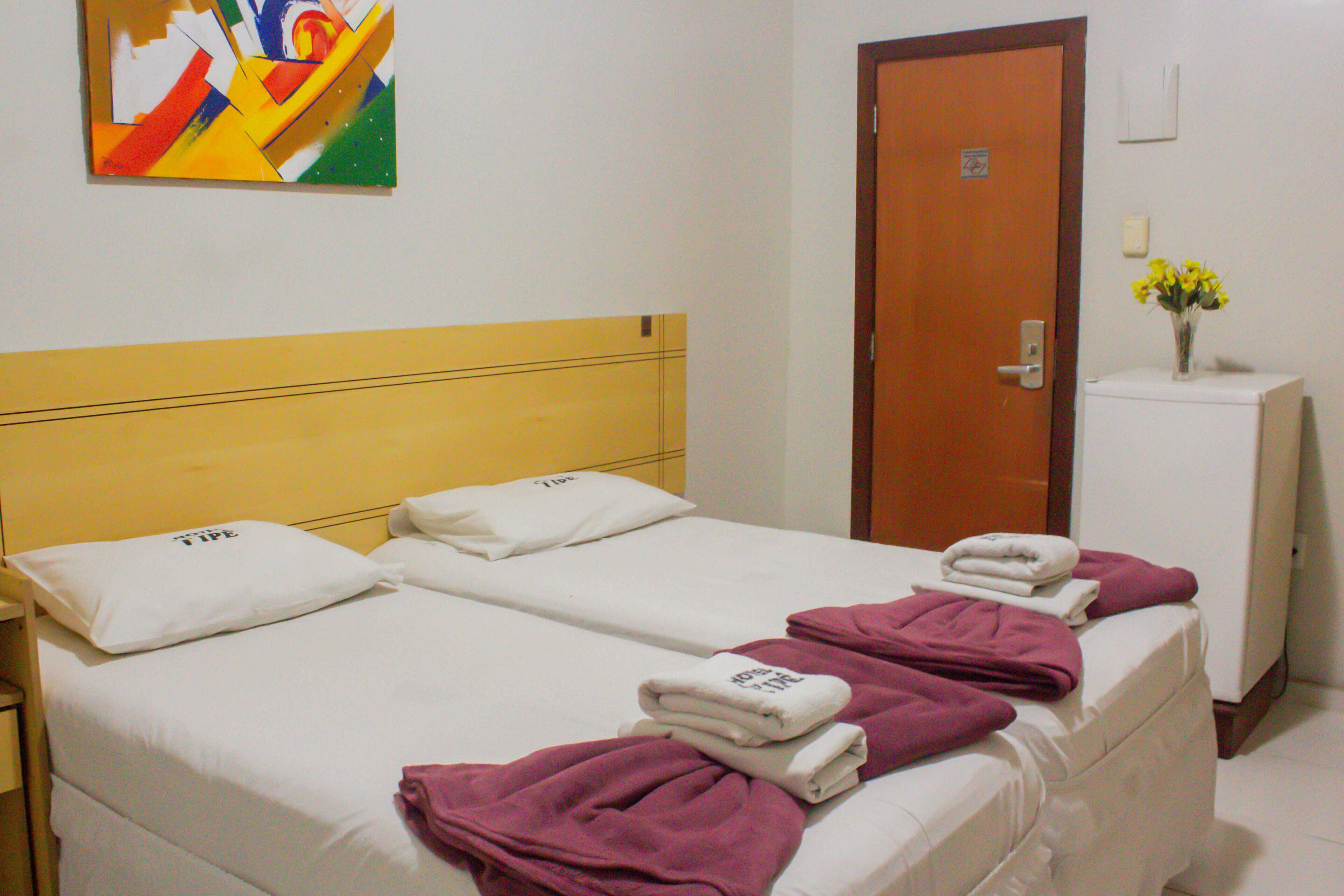 Quarto de hotel com duas camas, cada uma com roupa de cama e almofadas brancas, cabeceira de madeira, pequeno armário branco com um vaso de flores e um quadro abstrato colorido na parede.