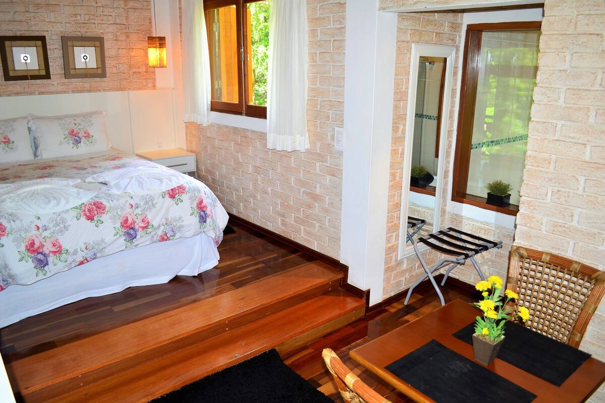 Quarto com piso de madeira, cama com roupa de cama floral, mesa de cabeceira, janela com cortinas, uma cadeira e uma mesa com planta em vaso.