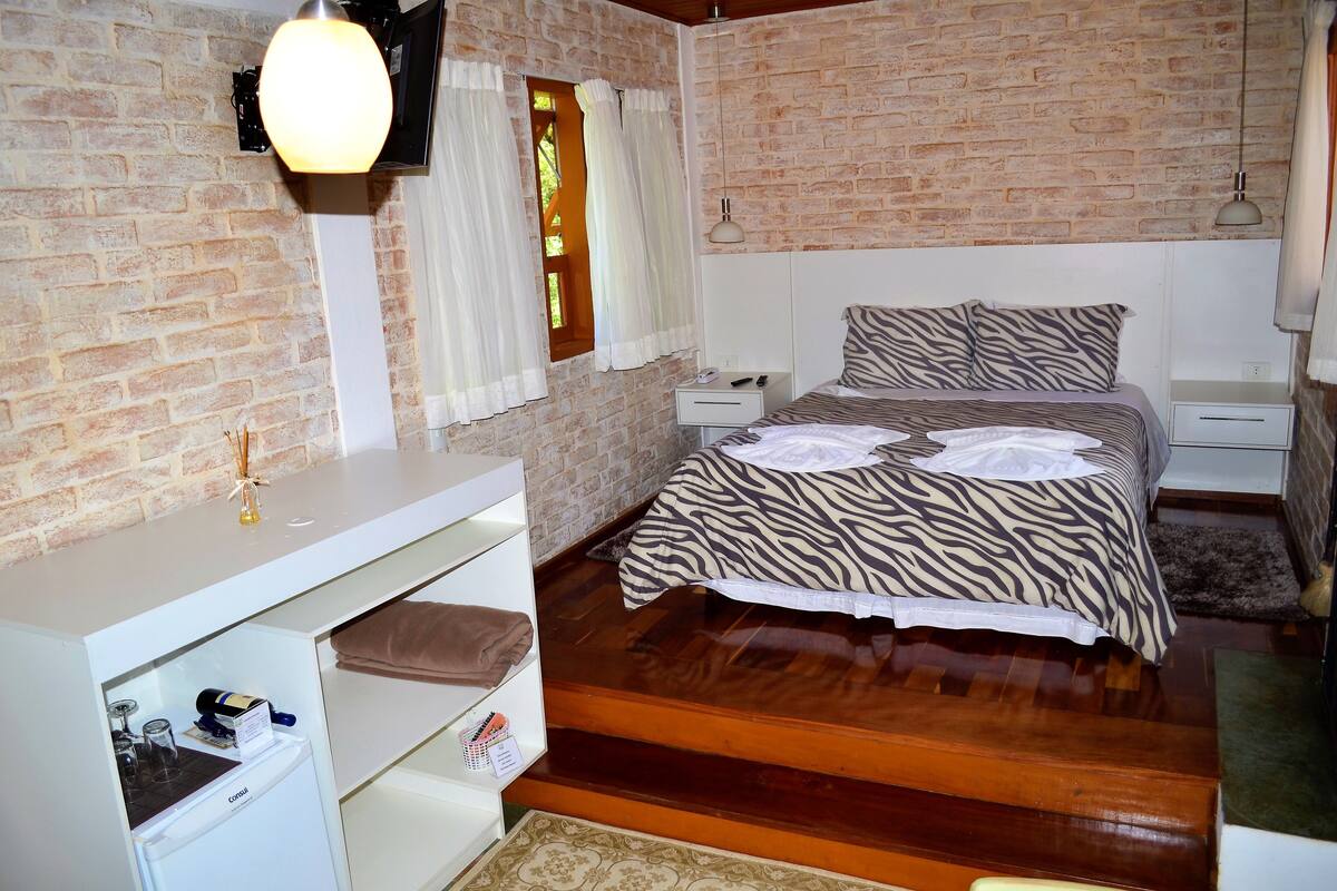 Quarto com parede de tijolos, cama de madeira, mesa de cabeceira branca, um criado-mudo, uma pequena escrivaninha e um mini-frigorífico embutido.
