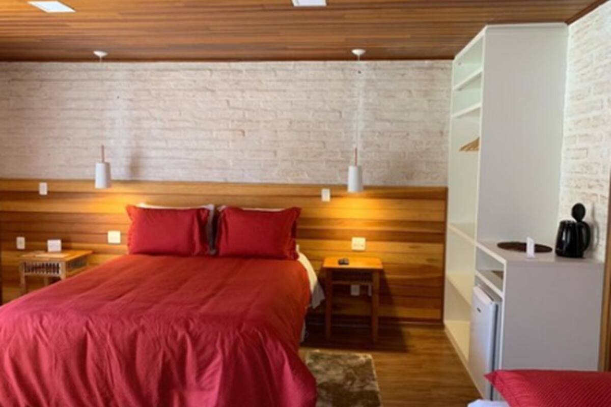 Quarto com teto de madeira, parede de tijolo, cama com roupa de cama vermelha, mesa de cabeceira, pequena mesa e guarda-roupa branco.