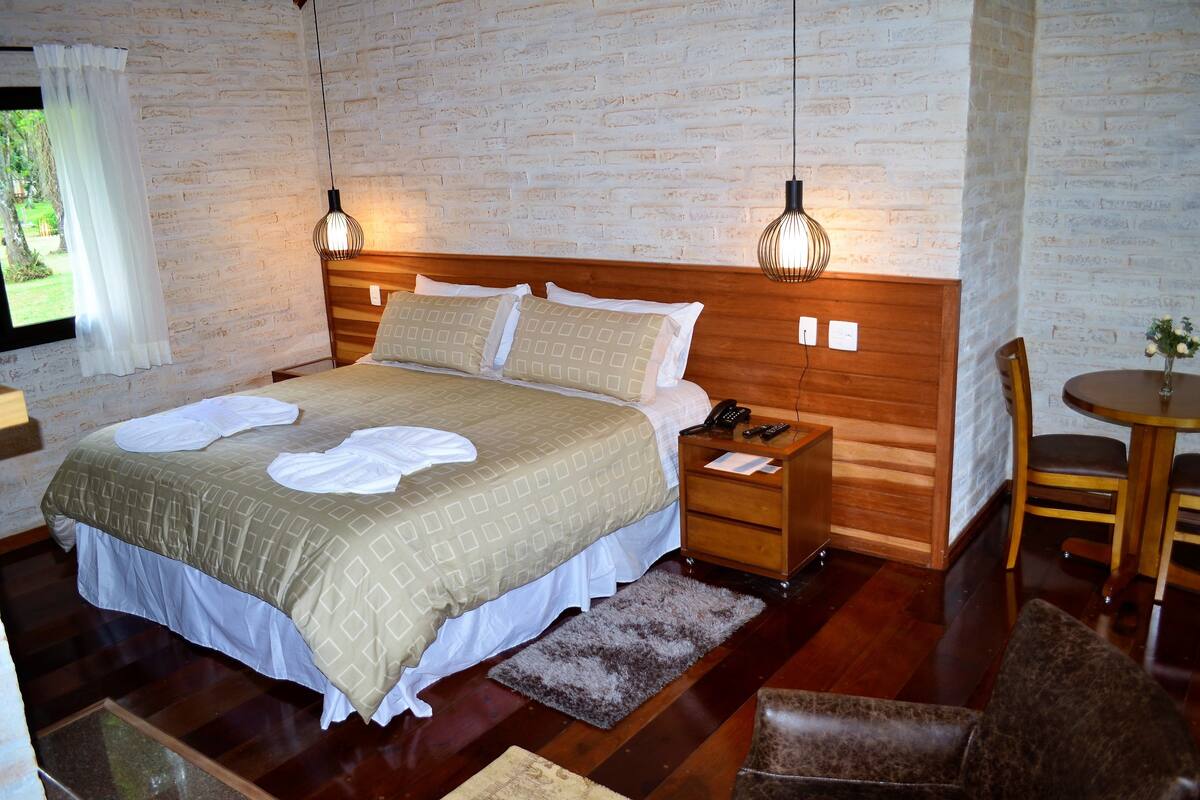Um quarto bem arrumado, com uma cama grande, mesas de cabeceira, cabeceira de madeira, uma mesa redonda com cadeiras e vista para o exterior pela janela.