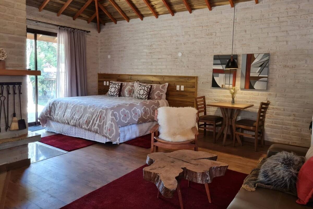 Quarto com cama grande, cabeceira de madeira, mesa de jantar com cadeiras e tapete vermelho.