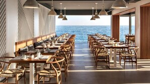 Ampla sala de jantar com mesas e cadeiras voltadas para uma grande janela com vista para o oceano.