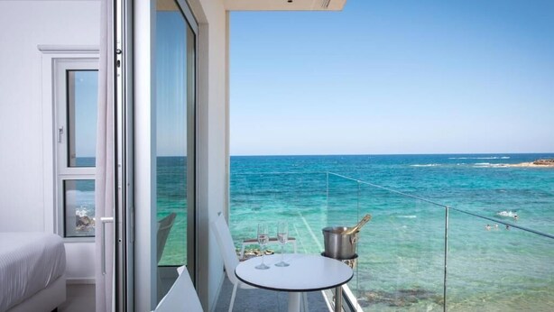 Uma varanda com vista desobstruída para o oceano, uma pequena mesa com uma garrafa e copos, e uma cadeira branca.