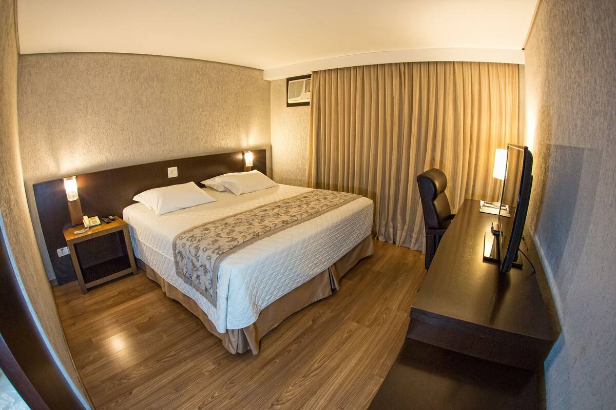 Quarto de hotel com uma cama grande, uma escrivaninha com cadeira, mesa de cabeceira e espelho.