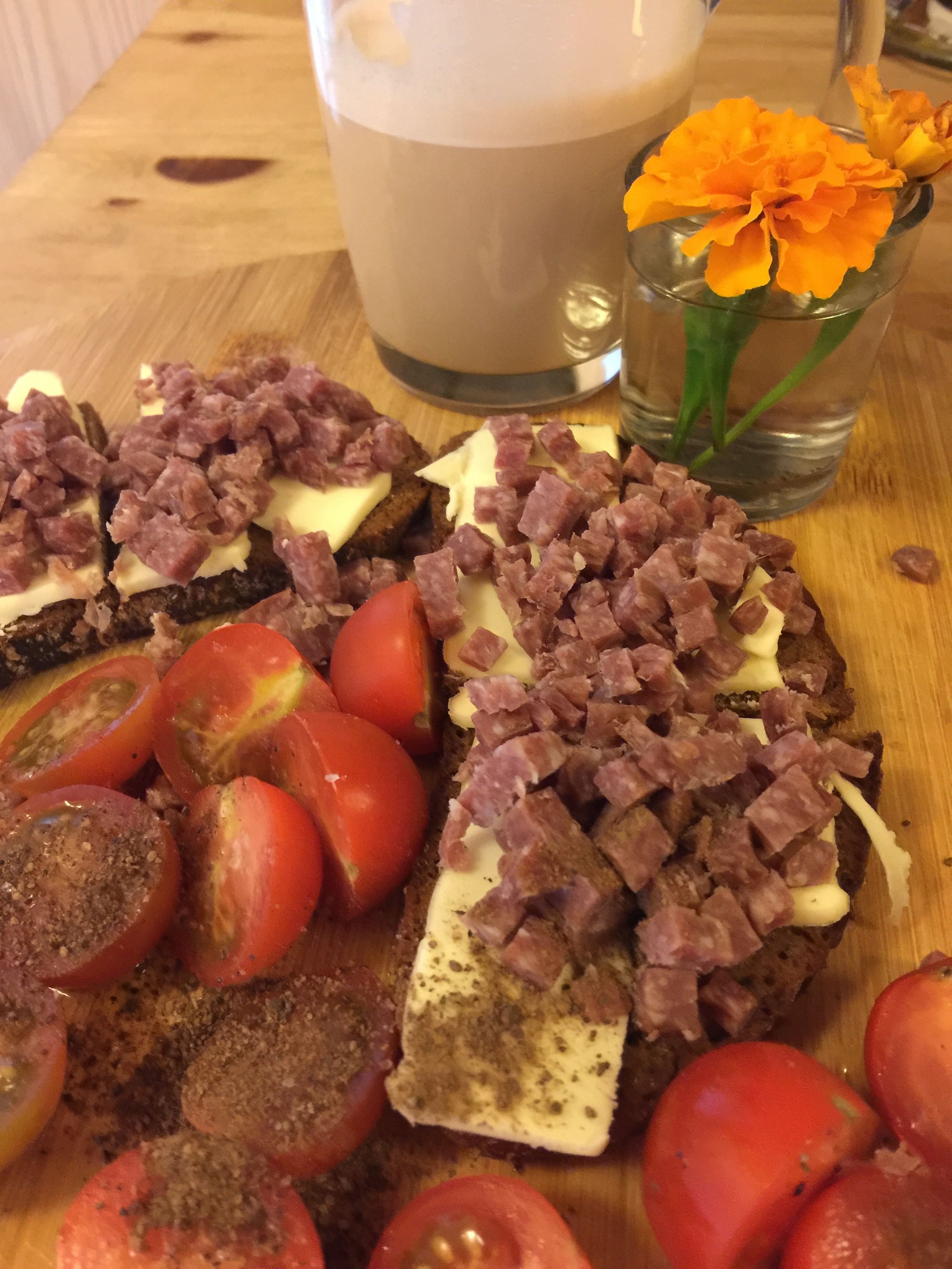 Uma mesa de madeira com uma refeição de pão coberto com queijo e fiambre, tomates e um copo de leite com uma flor.