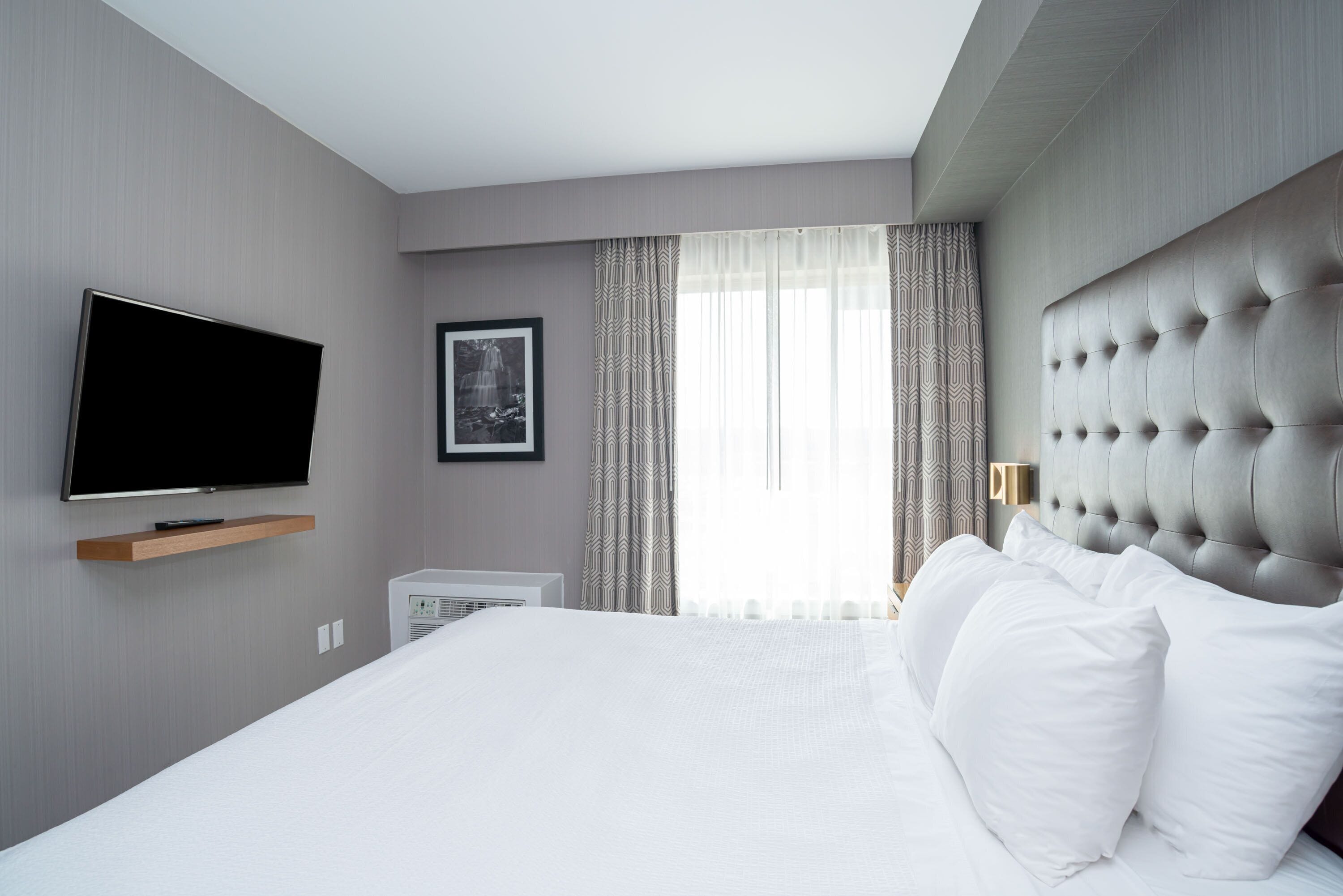 Quarto de hotel moderno com uma cama grande, televisão de tela plana na parede, um quadro pendurado na parede e uma janela com cortinas.