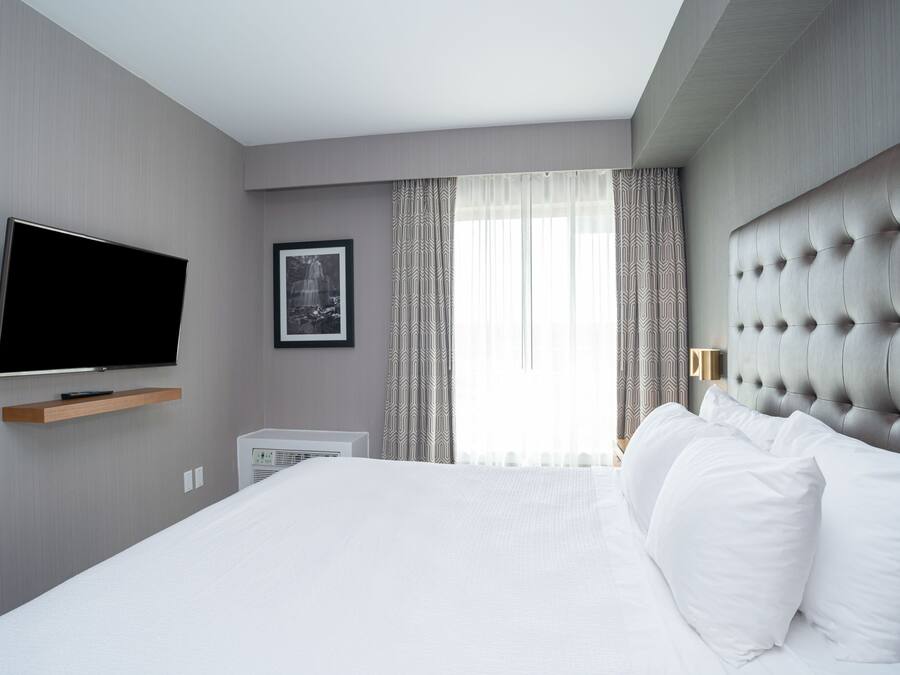 Quarto de hotel moderno com uma cama grande, televisão de tela plana na parede, um quadro pendurado na parede e uma janela com cortinas.