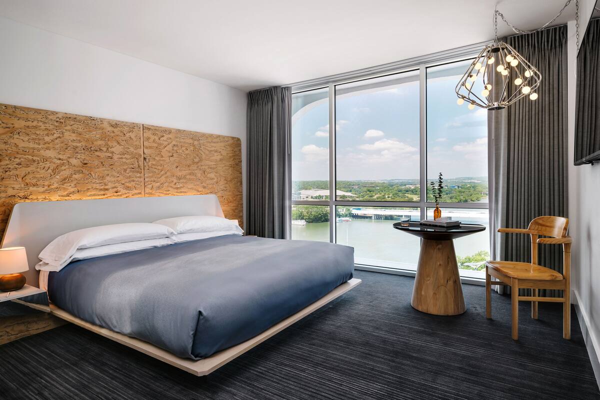 Quarto de hotel moderno com uma cama grande, uma mesa redonda de madeira e vista para a água através de grandes janelas.