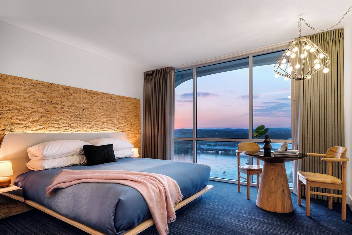 Quarto de hotel com uma cama grande, cabeceira de madeira, vista da cidade, área de refeições com mesa e cadeiras, e luminária pendente.