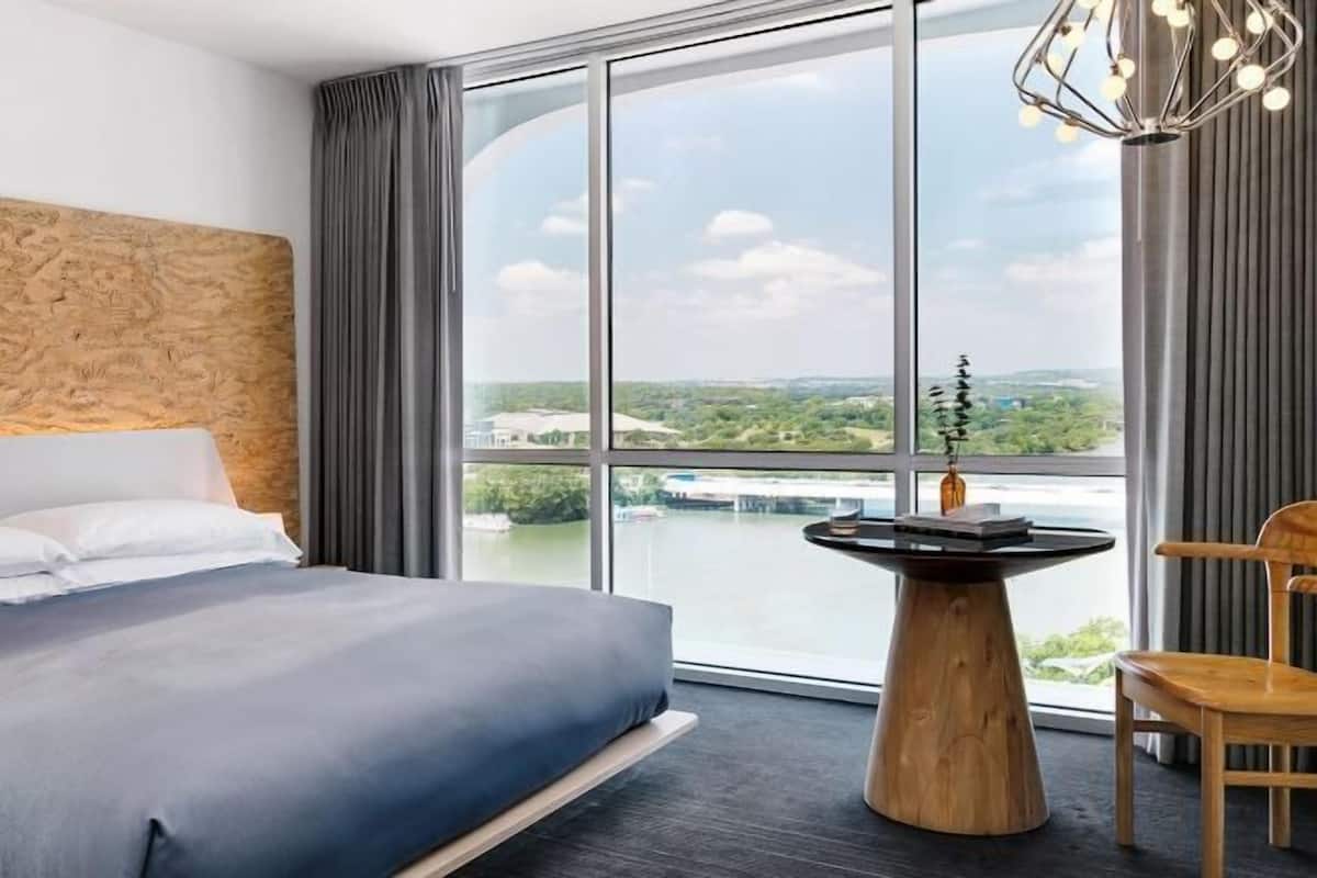 Quarto de hotel moderno com uma janela grande, uma cama, uma mesa de madeira redonda e uma cadeira.