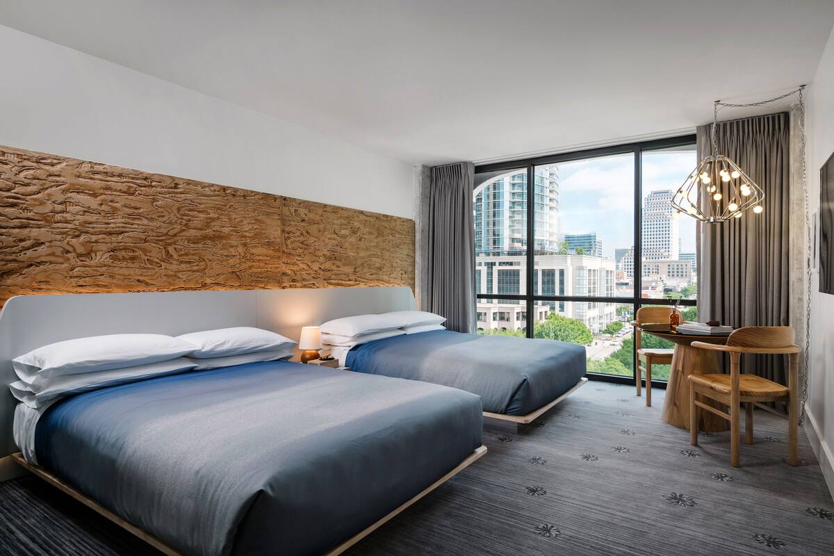 Quarto de hotel moderno com duas camas, cabeceira de madeira, vista para a cidade, área de jantar com mesa e cadeiras, e luminária pendente.