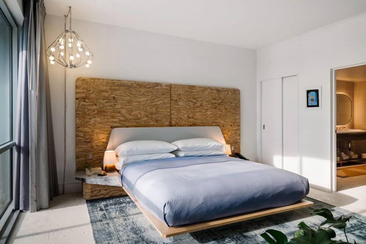 Quarto moderno com cabeceira de madeira, cama com colchão azul, mesa de cabeceira com abajur e uma janela grande com cortinas.