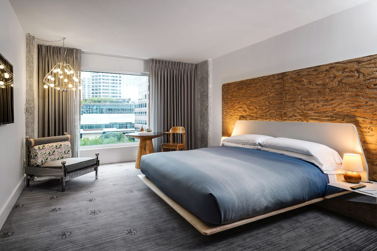 Quarto de hotel moderno com uma cama grande, uma mesa lateral com abajur, uma mesinha com cadeira e vista para a cidade pela janela.