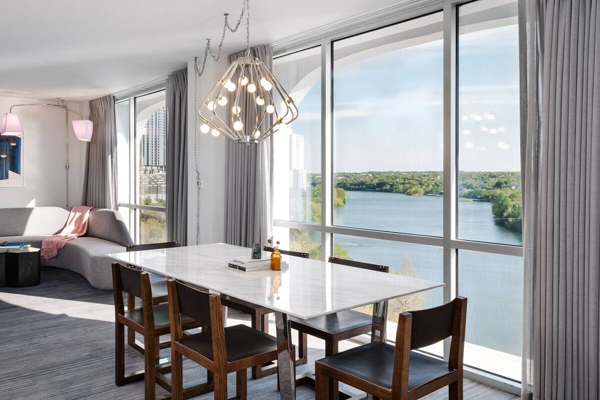 Quarto de hotel moderno com uma janela grande que oferece vista para um rio e edifícios. Há uma mesa de jantar com cadeiras, um sofá e um abajur.