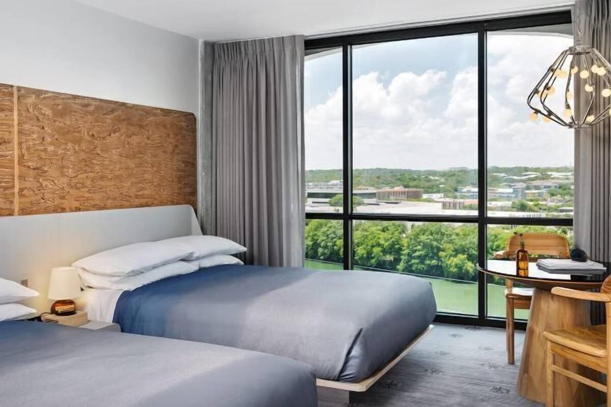 Quarto de hotel com duas camas, uma janela grande com vista para a cidade, cabeceira de madeira e uma pequena mesa com uma cadeira.