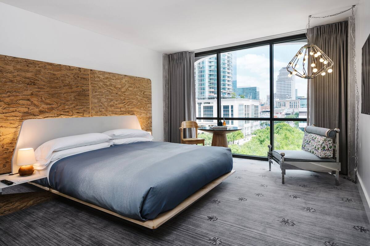 Quarto de hotel moderno com uma cama grande, uma mesa pequena, uma cadeira e vista da cidade por uma janela ampla.