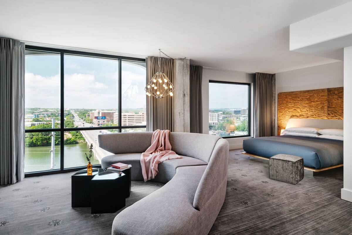 Quarto de hotel moderno com uma grande janela, um sofá, uma cama e uma pequena mesa.
