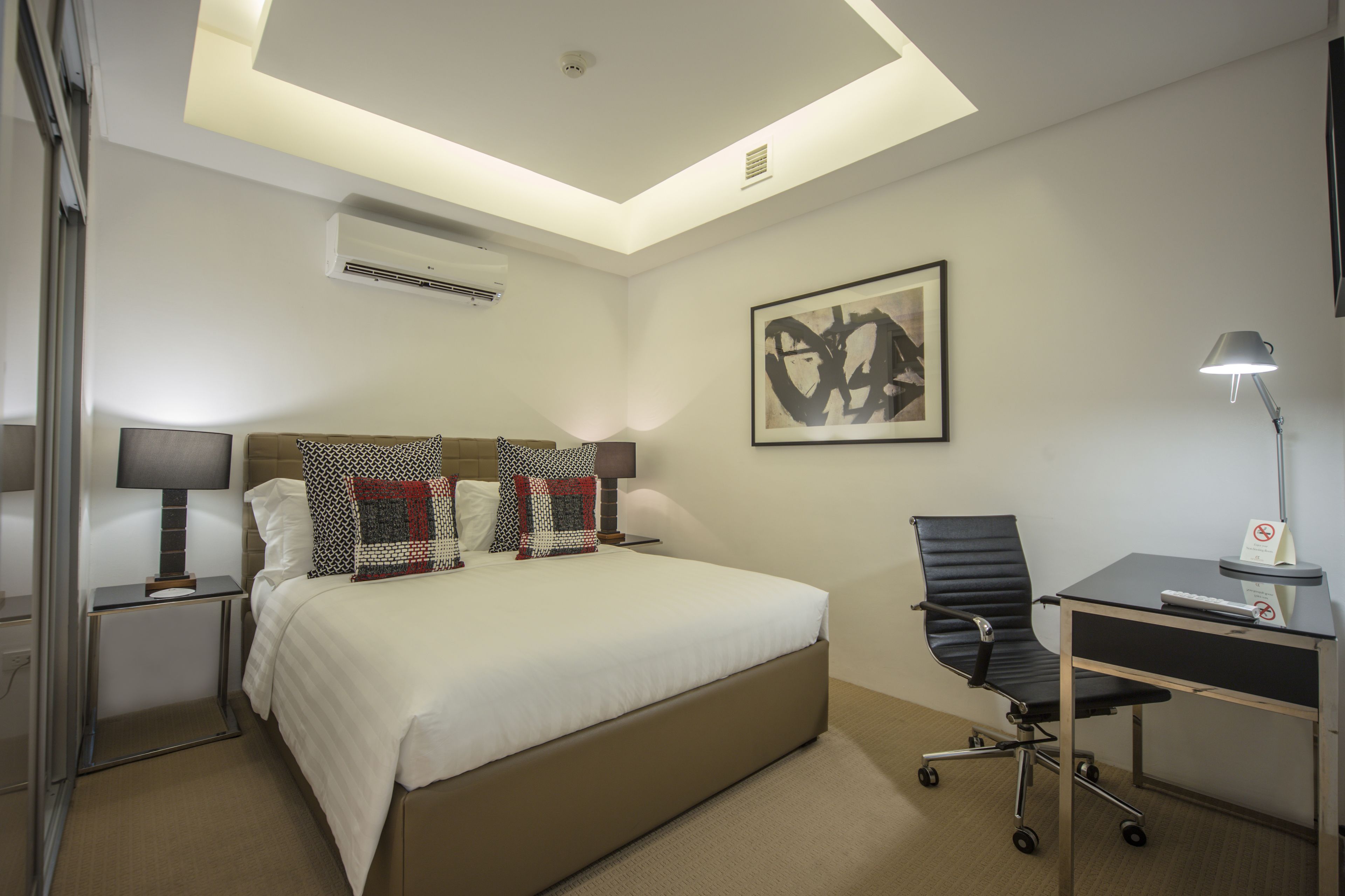Quarto de hotel com uma cama grande, uma escrivaninha com cadeira, uma luminária e um quadro abstrato na parede.