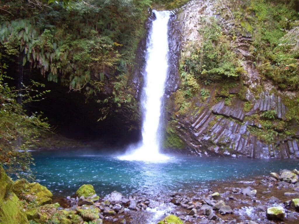 Uma cascata deságua em uma piscina azul límpida, rodeada por terreno rochoso e vegetação densa.