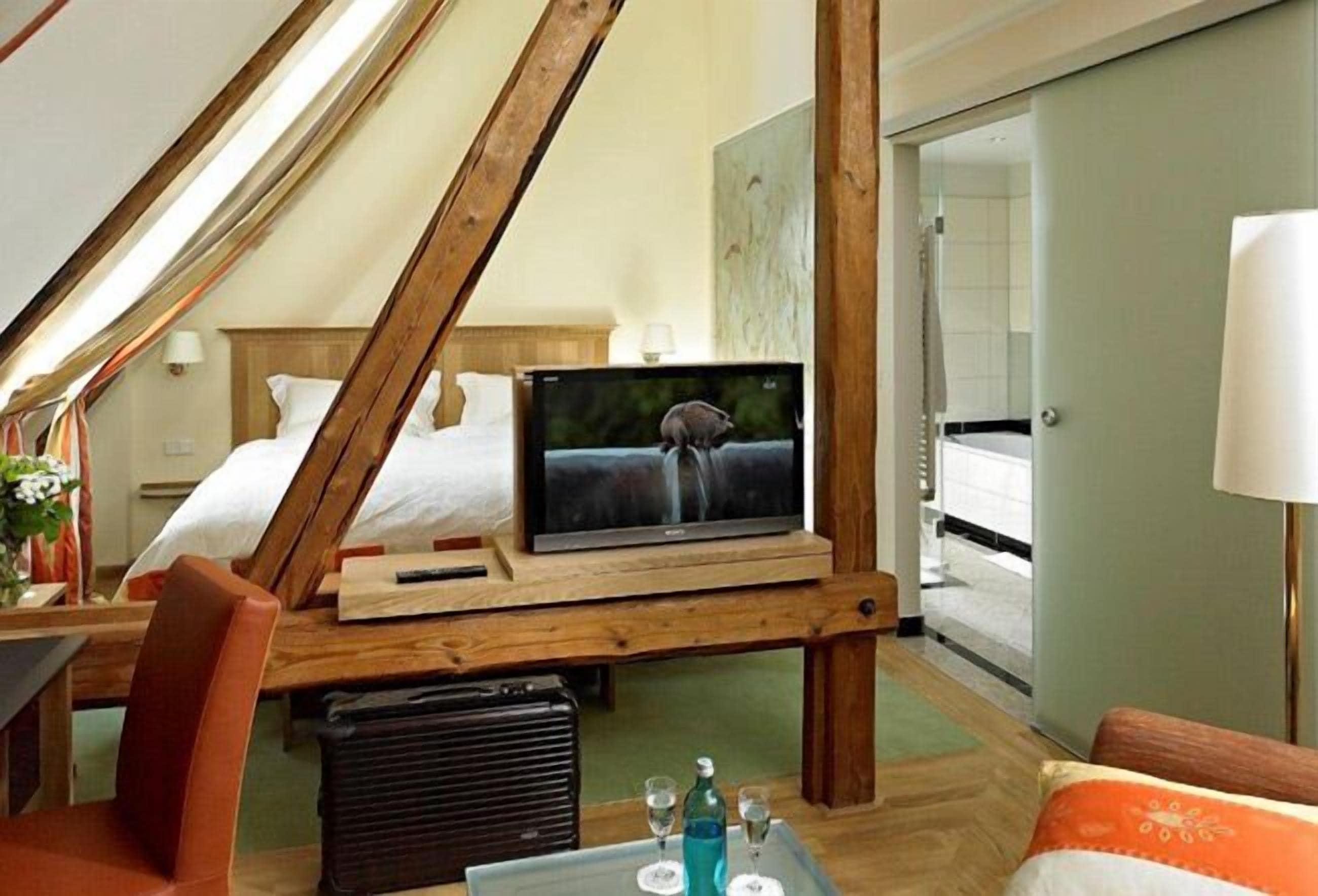 Quarto de hotel com cama, televisão sobre suporte de madeira, escrivaninha com cadeira e mesa com copos e garrafas.