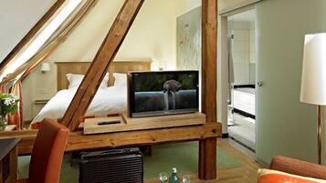Quarto de hotel com cama, televisão sobre suporte de madeira, escrivaninha com cadeira e mesa com copos e garrafas.