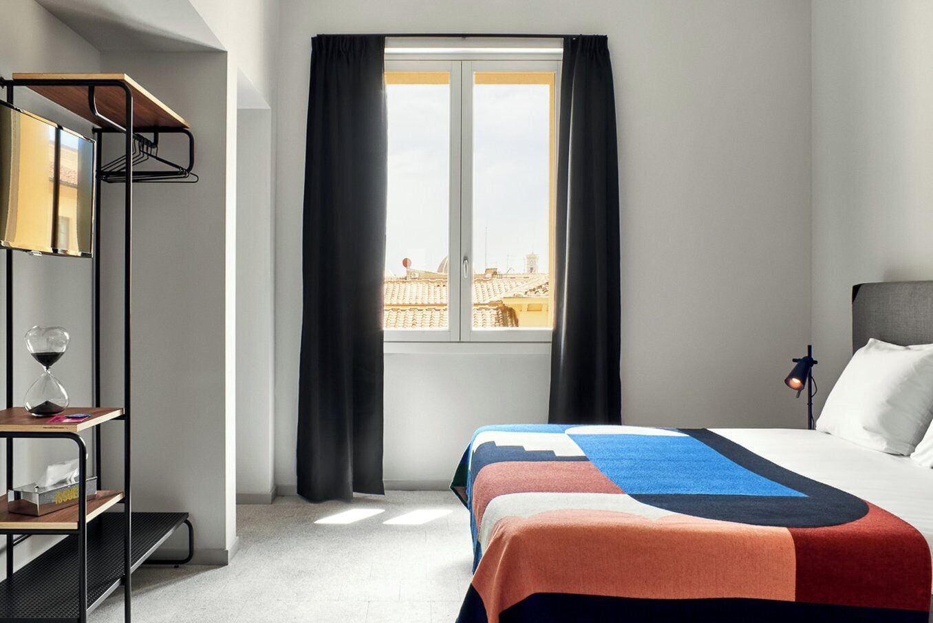 Quarto de hotel com cama, escrivaninha com cadeira, janela com cortinas e vista para prédios.