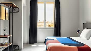 Quarto de hotel com cama, escrivaninha com cadeira, janela com cortinas e vista para prédios.