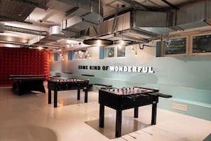 Um quarto com duas mesas de futebol de mesa, um painel acústico vermelho e uma parede com a frase 'SOME KIND OF WONDERFUL'.