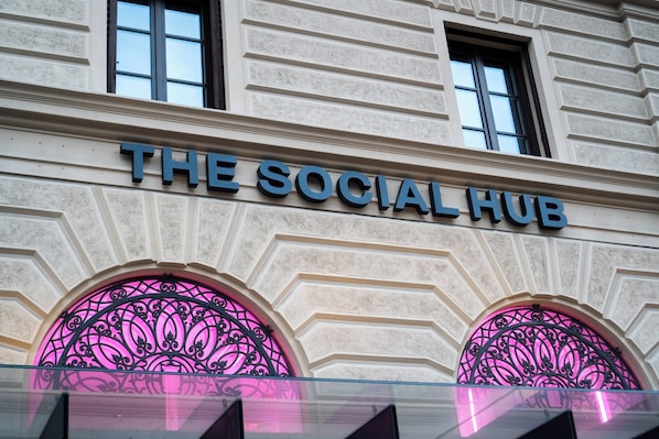 Fachada de edifício com o letreiro 'THE SOCIAL HUB' acima de duas janelas em arco com detalhes em ferro forjado.