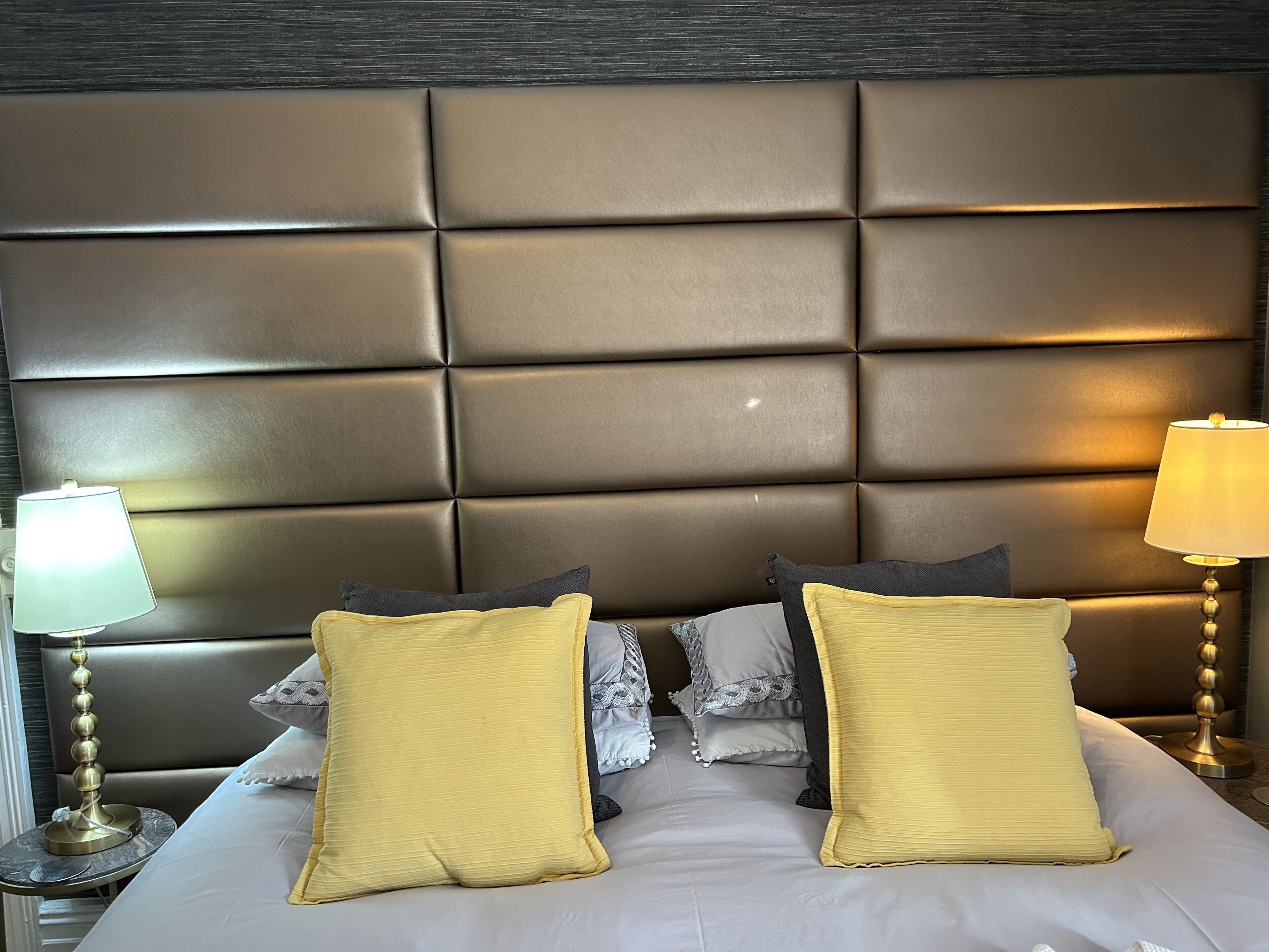 Quarto de hotel com cabeceira acolchoada, duas luminárias de cabeceira e várias almofadas.