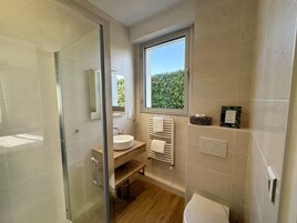 Casa de banho moderna com cabine de duche em vidro, lavatório com espelho, janela com vista para vegetação e sanita.