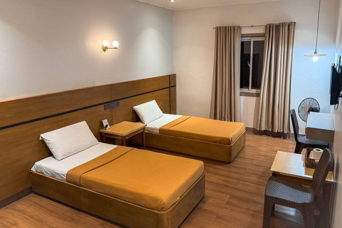 Quarto de hotel com duas camas, uma mesa, uma cadeira, um ventilador de teto e uma janela com cortinas.