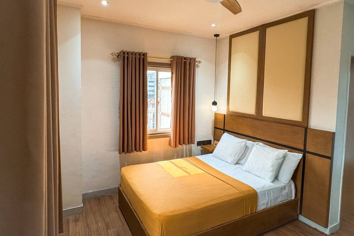 Quarto de hotel com cama, ventoinha no teto, janela com cortinas e mesa de cabeceira.