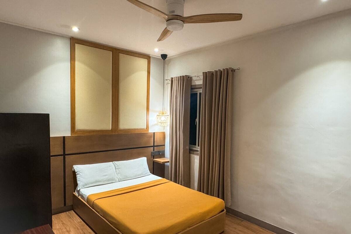 Quarto de hotel com cama, painel de cabeceira de madeira, ventilador de teto e janela com cortinas.