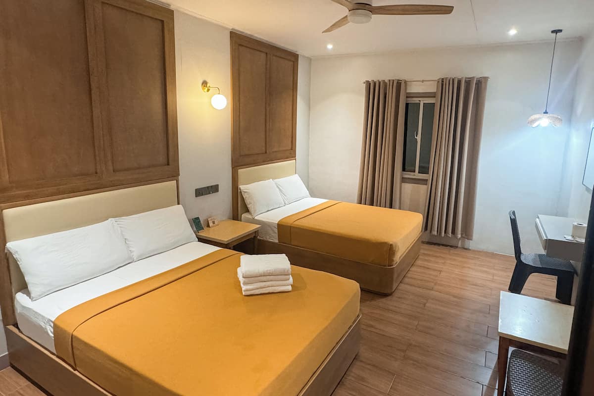 Quarto de hotel com duas camas, um ventilador de teto, uma escrivaninha e uma janela com cortinas.