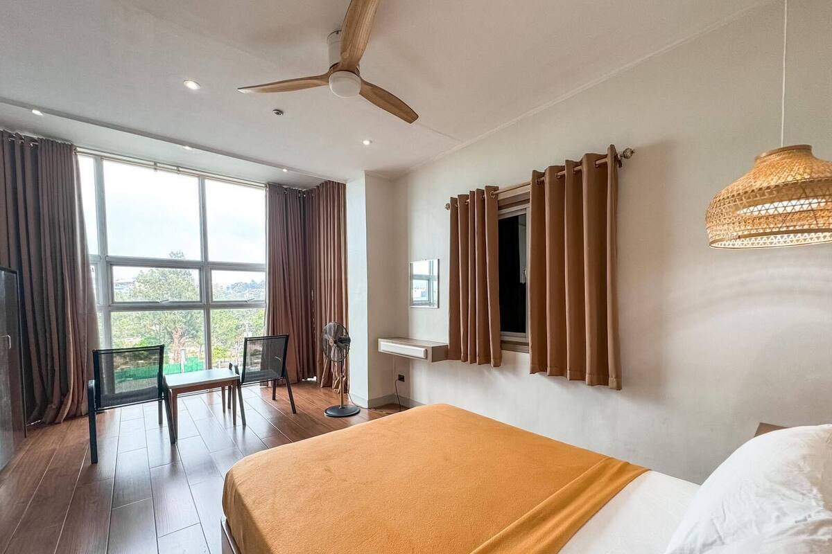 Quarto de hotel com cama, escrivaninha, cadeira, ventilador de teto e janela com cortinas.