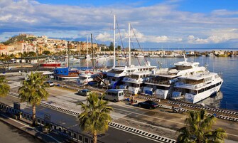 Uma marina com vários barcos atracados, uma estrada com veículos e edifícios ao fundo.
